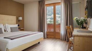 Quarto de hotel com cama, escrivaninha, cadeira, janela com cortinas e vista para uma área arborizada.