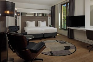 Quarto de hotel com cama, escrivaninha, cadeira, televisão e janela com cortinas.