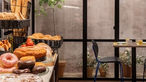 Vitrine de padaria com diversos produtos de panificação, uma janela com vista para o verde e uma mesa com uma cadeira.