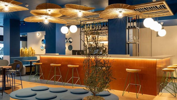 Uma área de bar moderna com balcão de madeira, banquetas e um espaço de estar central com almofadas redondas.