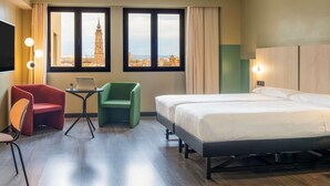Quarto de hotel com cama, escrivaninha com um laptop, uma cadeira vermelha, uma cadeira verde e vista para uma torre por grandes janelas.