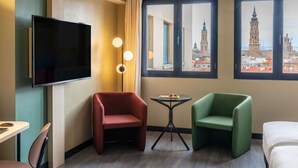 Quarto de hotel com televisão, duas cadeiras, uma mesinha e uma cama.