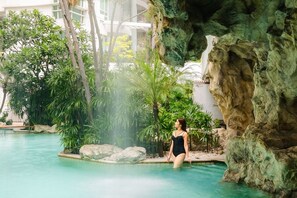 Mulher de fato de banho preto em frente a piscina, com uma gruta rochosa ao fundo.