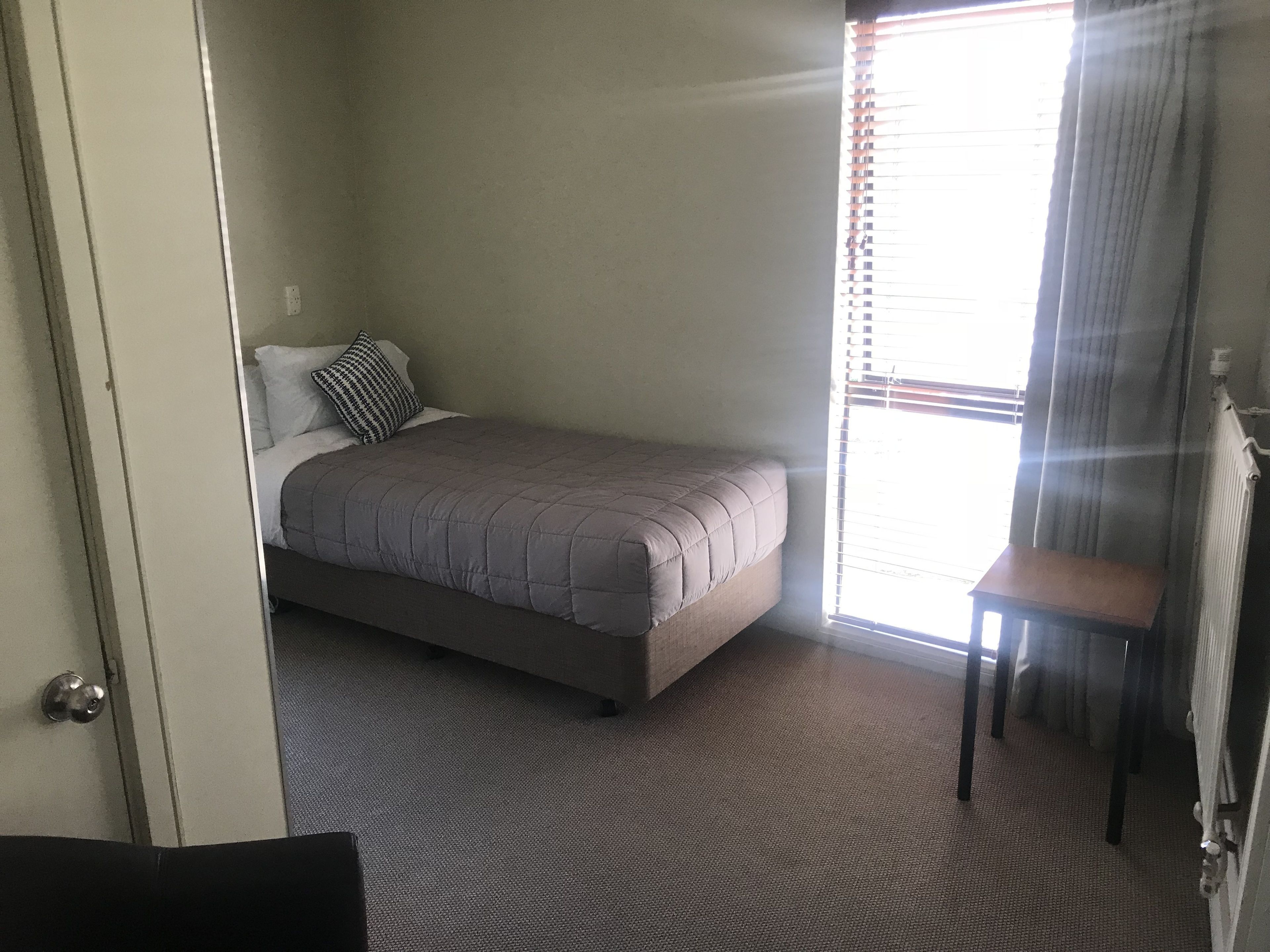 Quarto com cama, uma mesinha e janela com persianas.