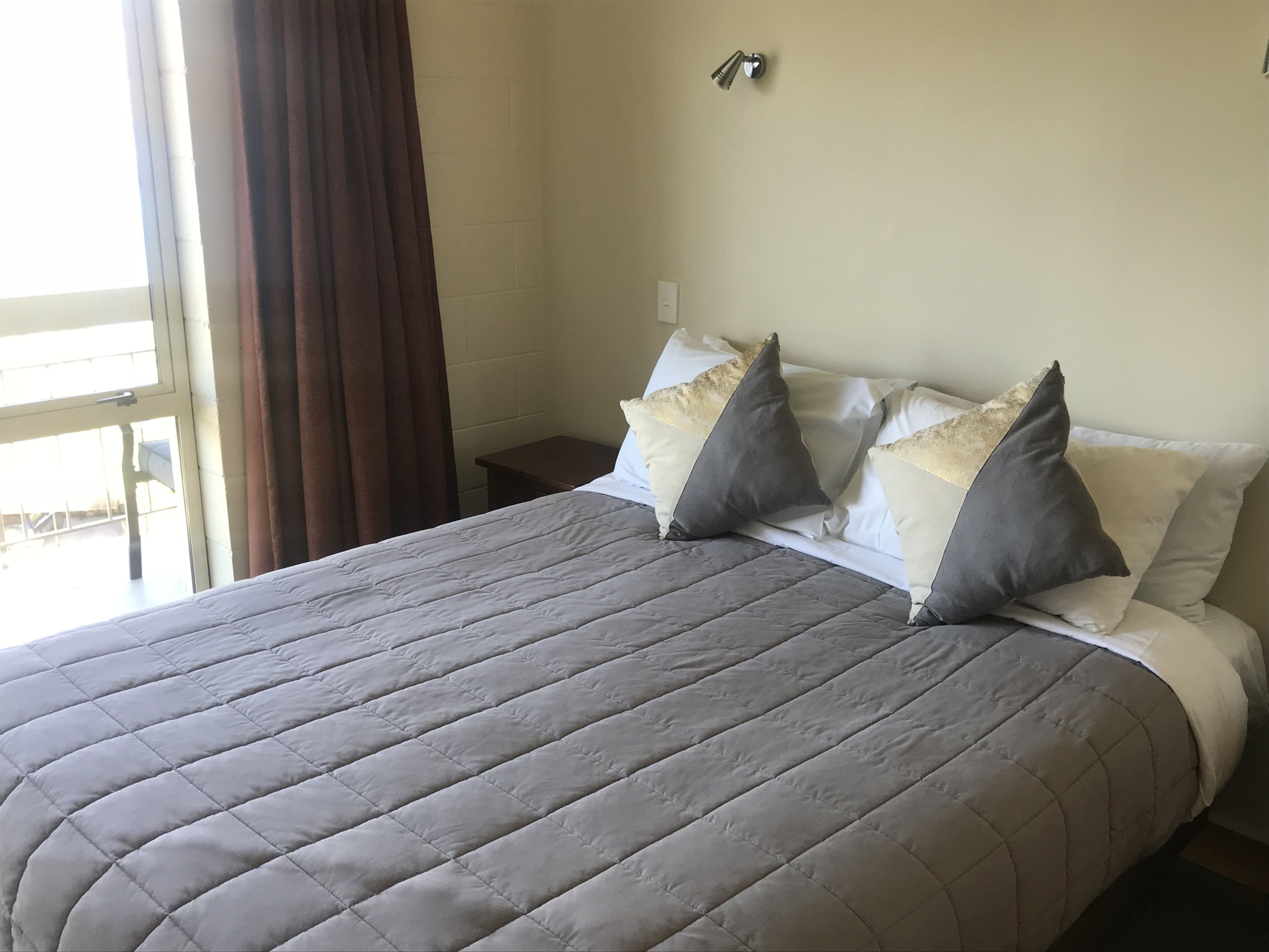 Cama bem arrumada com um edredom cinzento, duas almofadas e uma mesinha de cabeceira.