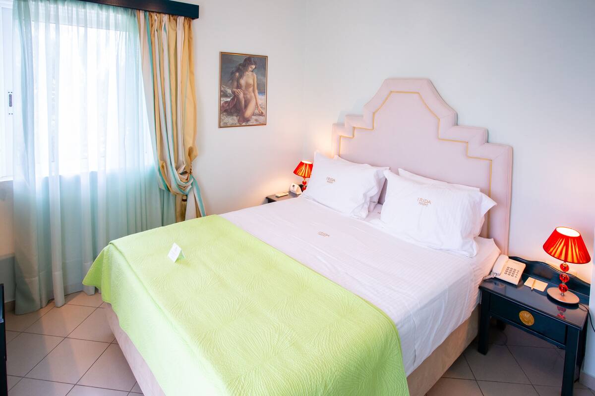 Quarto de hotel com cama, mesa de cabeceira com abajur, quadro na parede e janela com cortinas.