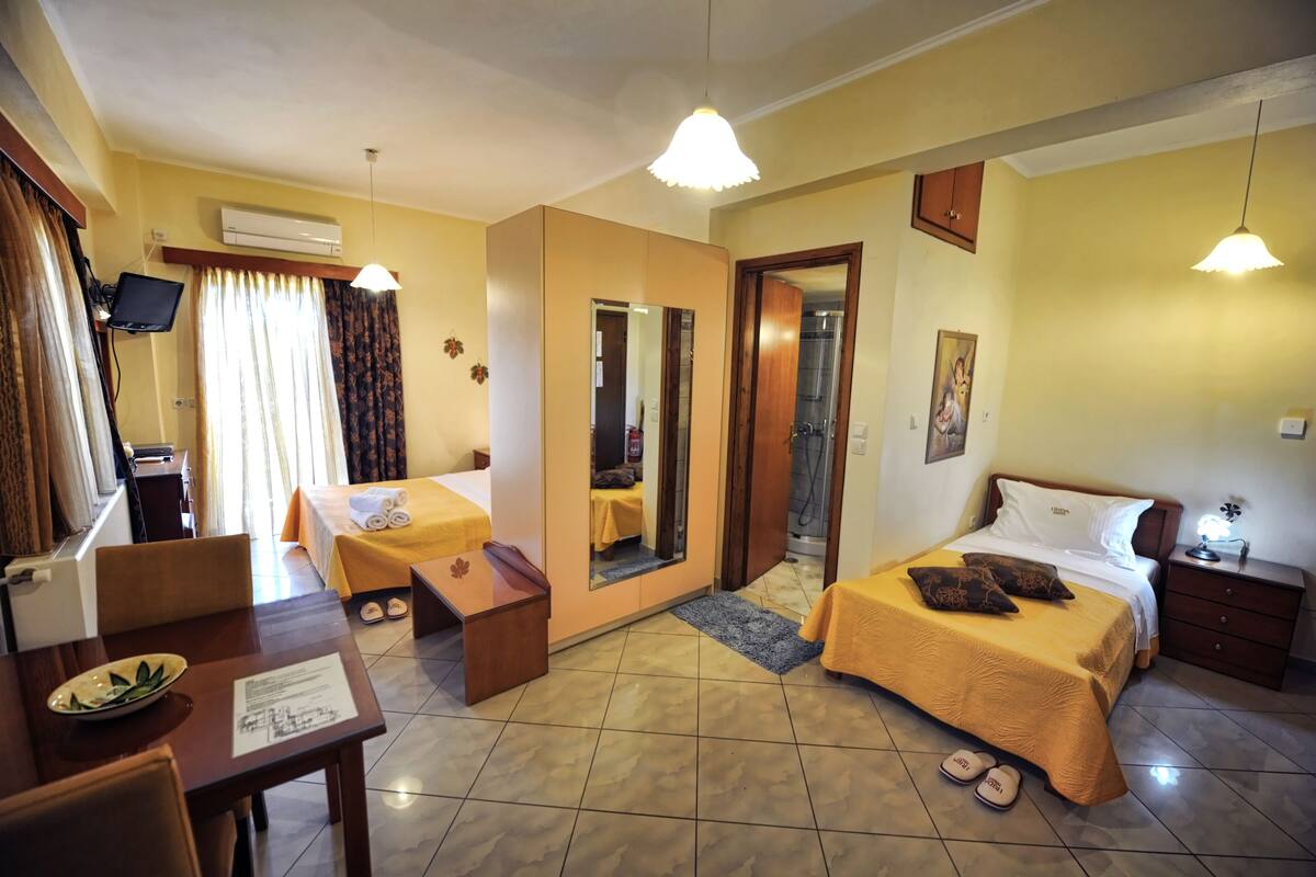 Quarto de hotel com duas camas, uma escrivaninha, uma cadeira, um espelho, um guarda-roupa e uma mesinha de cabeceira com chinelos.