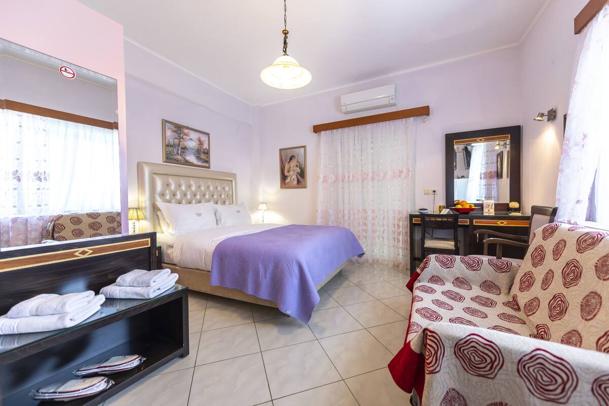 Quarto de hotel com cama, escrivaninha, cadeira, prateleira com toalhas e espelho.