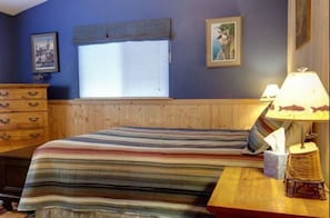 Quarto com cama, cómoda de madeira, janela com persianas e uma luminária.
