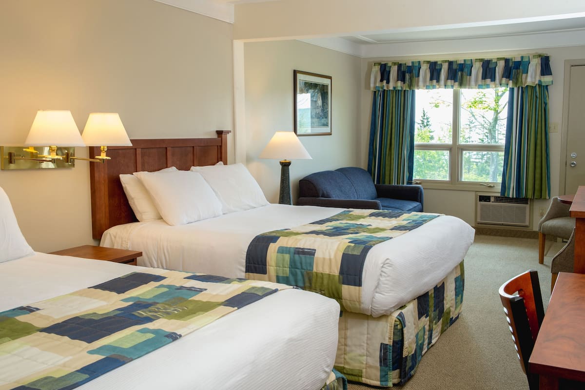 Quarto de hotel com duas camas, uma mesa de trabalho, uma cadeira, janela com cortinas e luminárias fixadas na parede.