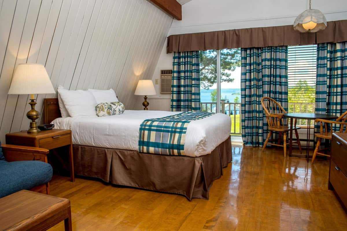 Quarto com cama, abajures, cômoda de madeira, cadeira e vista para o exterior.