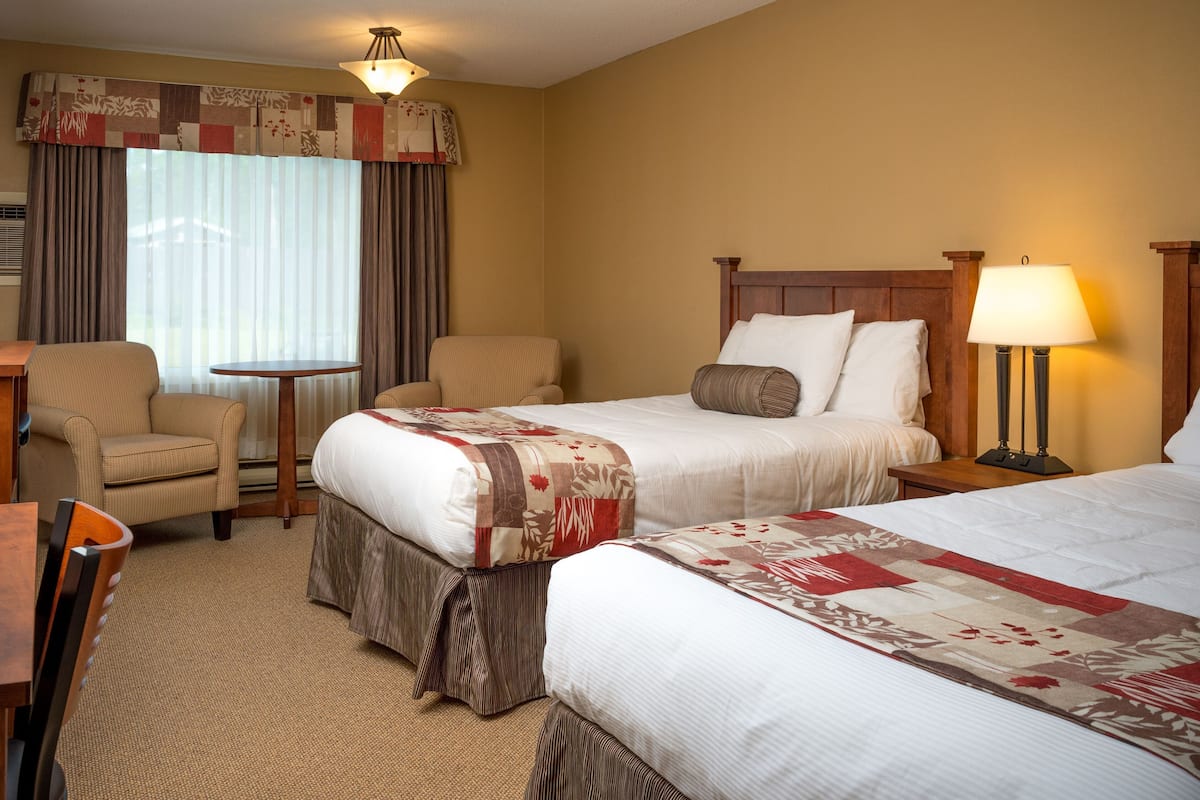Quarto de hotel com duas camas, uma mesinha, uma cadeira e um abajur.