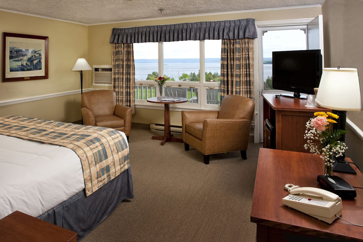 Quarto de hotel com cama, duas poltronas, uma mesinha, secretária com telefone, televisão e janela com vista para o mar.
