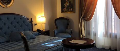 Quarto de hotel com uma cama grande, duas poltronas, uma mesa pequena, um abajur e um quadro na parede.