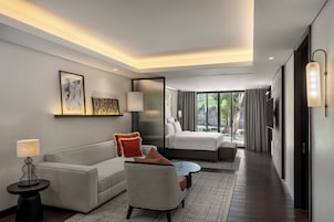 Quarto de hotel moderno com sofá, poltrona, mesa de cabeceira, luminária e uma cama com vista para o exterior.