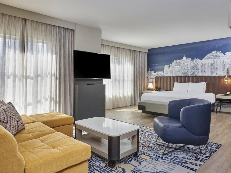 Quarto de hotel com cama, sofá, televisão e um mural com vista para a cidade.