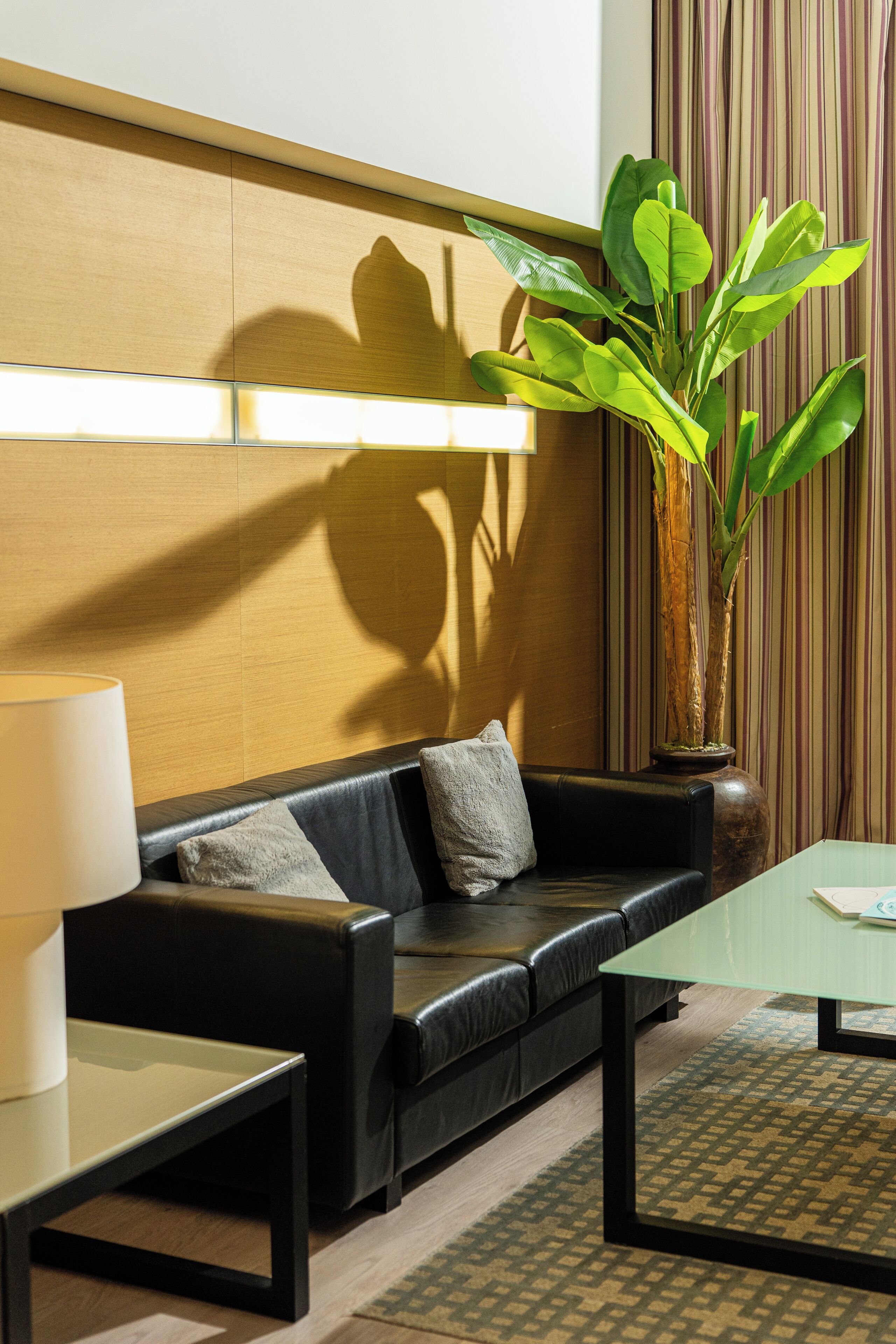 Quarto de hotel moderno com sofá de couro preto, mesa de centro de vidro, luminária, planta em vaso e cortina listrada.