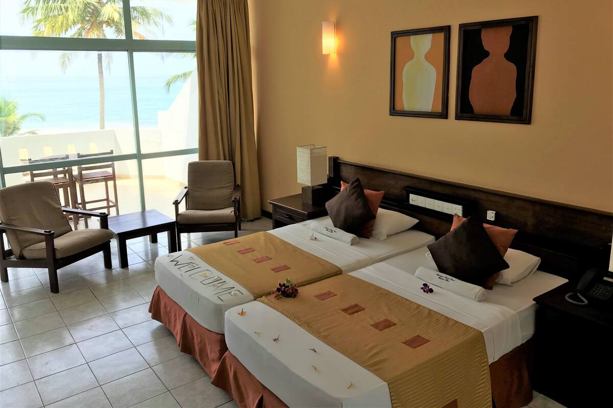 Quarto de hotel com duas camas, uma escrivaninha, uma cadeira e vista para o oceano.