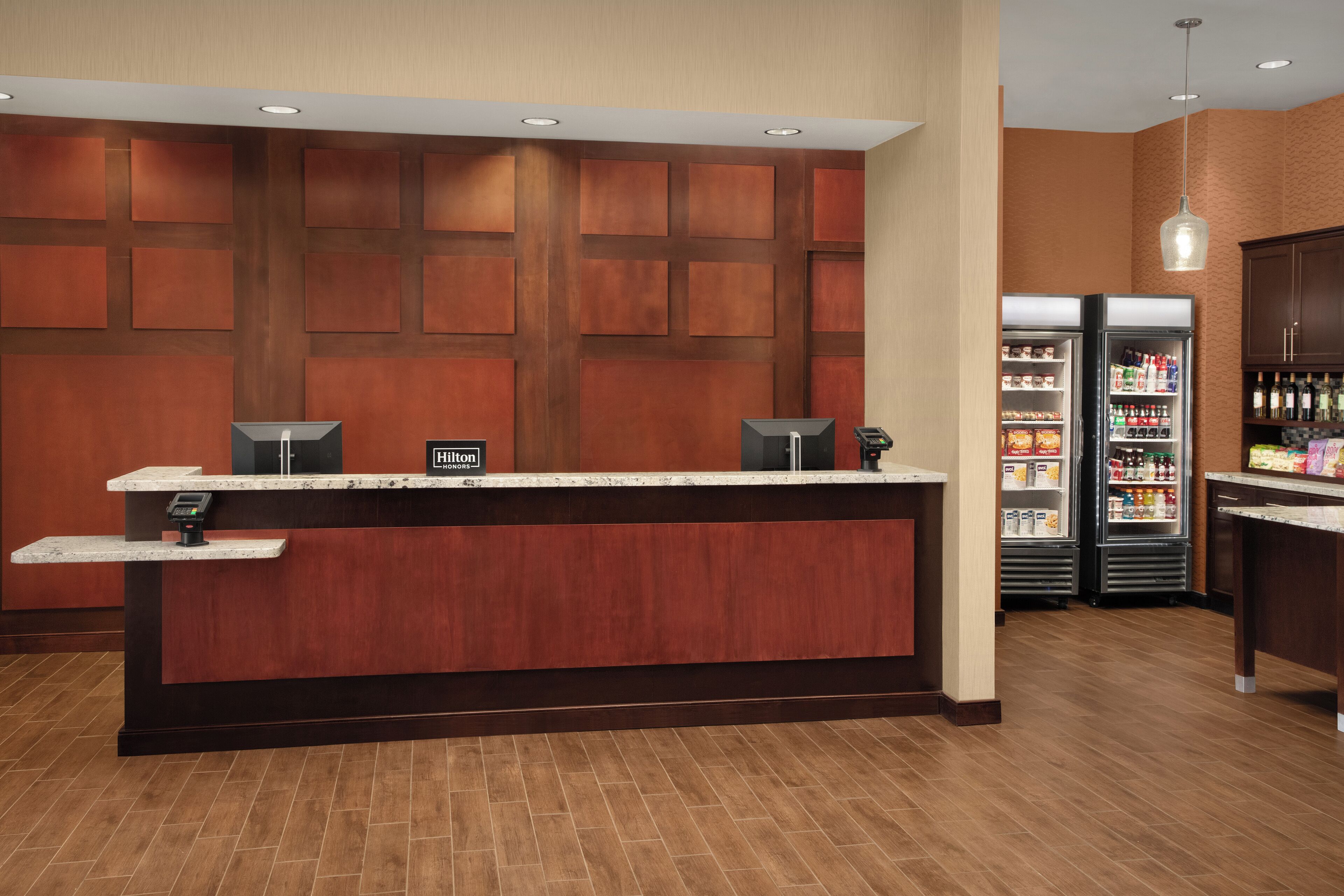 Área de recepção de hotel com parede de painéis de madeira, balcão de recepção, dois refrigeradores abastecidos com bebidas e um lustre.