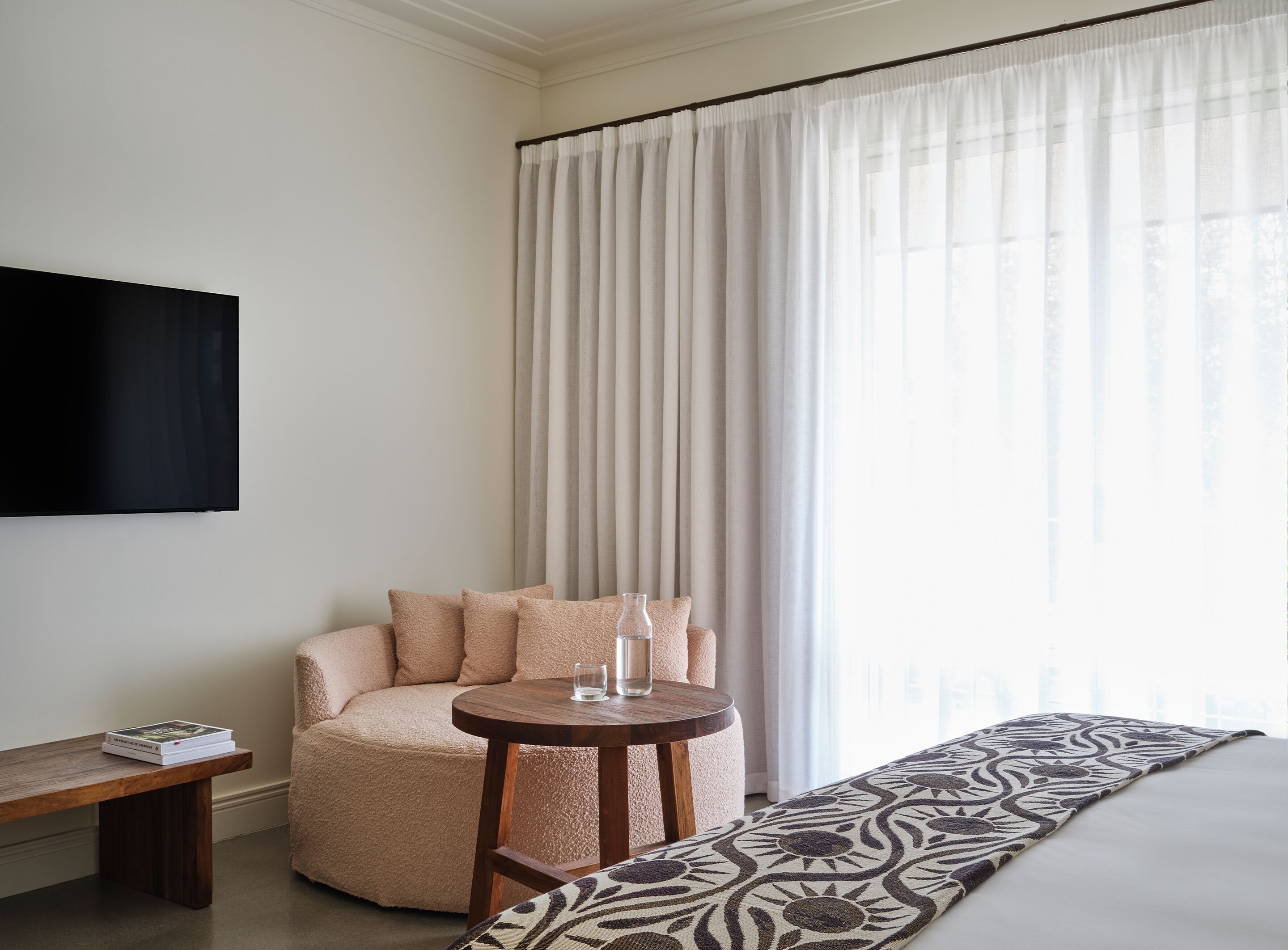Quarto de hotel moderno com uma cama grande, uma mesa de madeira redonda, um sofá com almofadas, uma TV de tela plana na parede e cortinas translúcidas na janela.