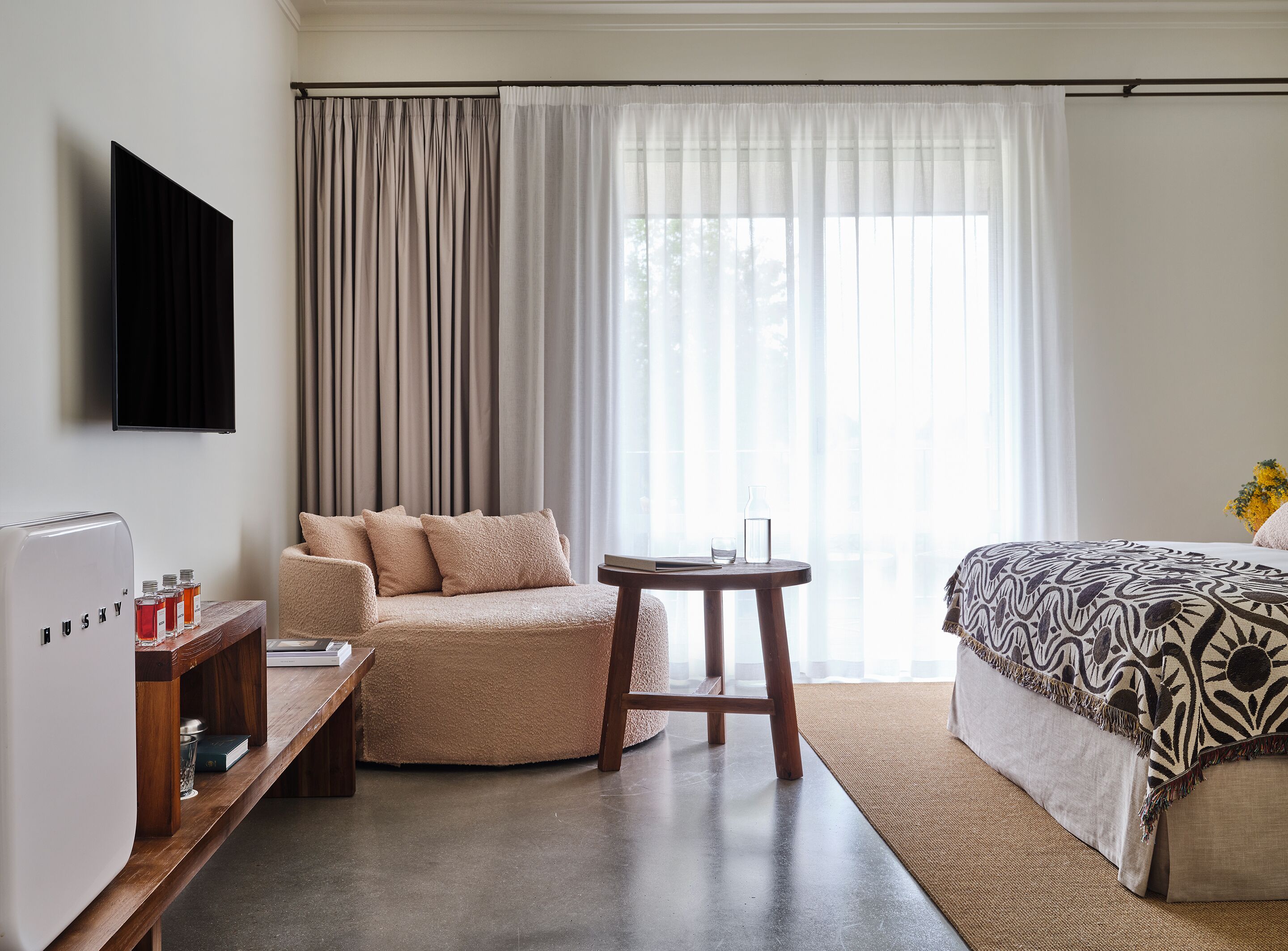 Quarto de hotel moderno com sofá, uma pequena mesa, cama com roupa de cama estampada, televisão de tela plana e minibar.