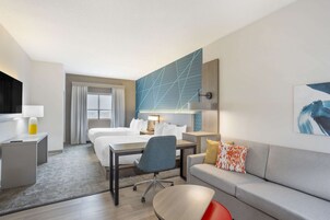 Quarto de hotel com cama, escrivaninha, cadeira, sofá e uma janela grande.