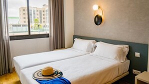 Quarto de hotel com duas camas, janela com cortinas, luminária fixada na parede e vista para edifícios.
