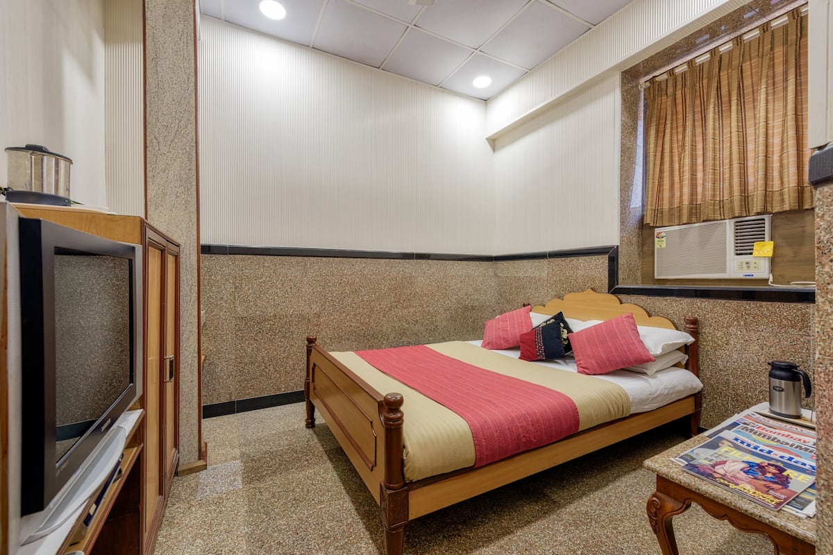 Quarto de hotel com cama de madeira, televisão, uma mesa pequena com uma revista e janela com cortinas.
