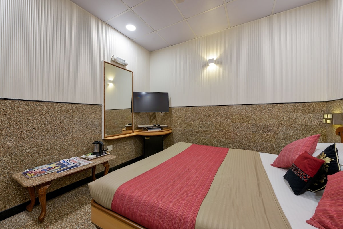 Quarto de hotel com cama, escrivaninha com televisão de tela plana, espelho e pequena mesa com revista e cafeteira.