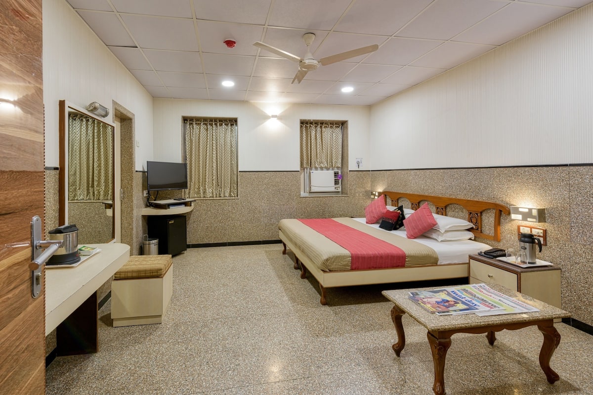 Quarto de hotel com uma cama, mesa de cabeceira, escrivaninha com cadeira, televisão e ventoinha no teto.