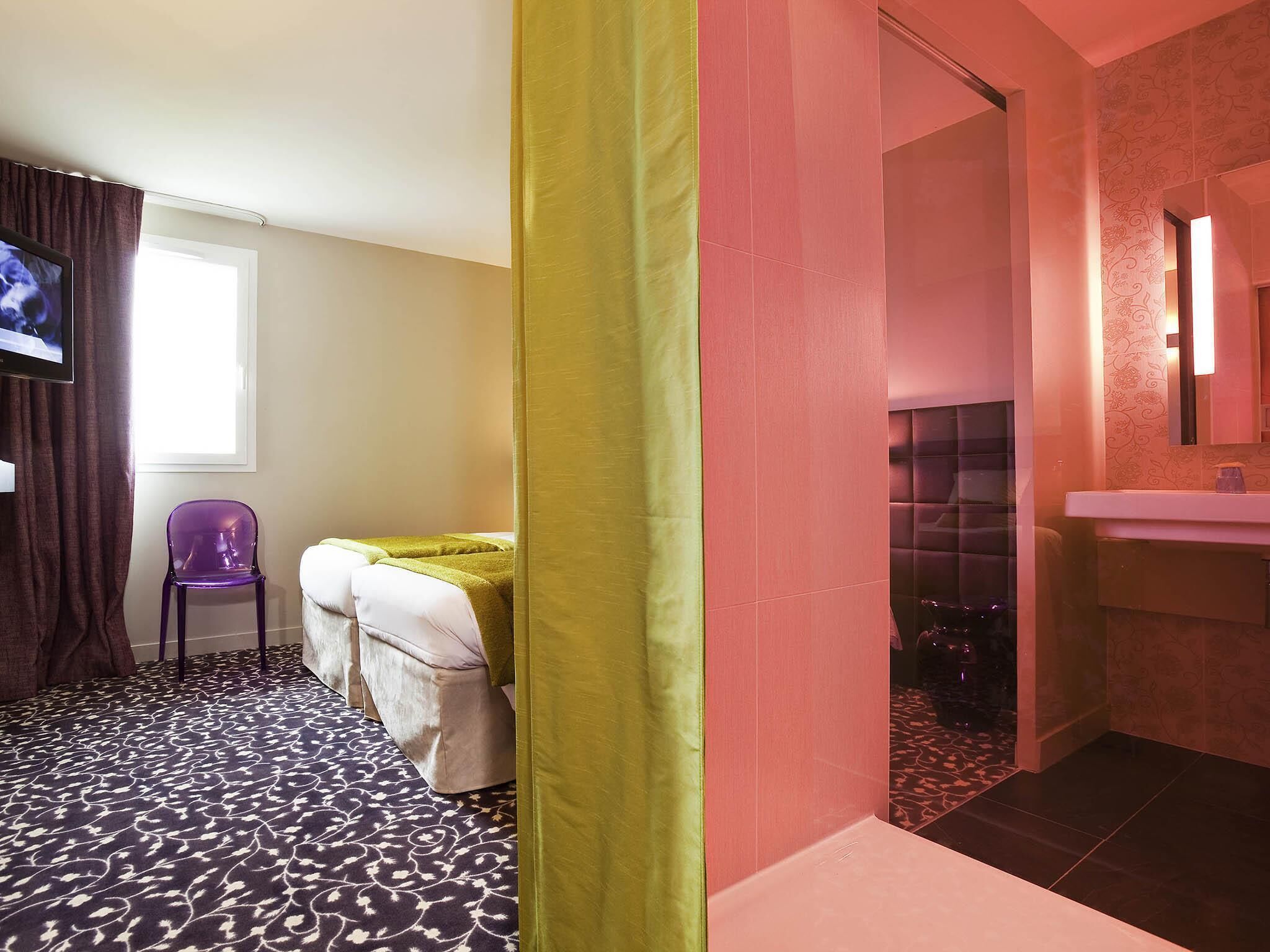 Quarto de hotel com cama, televisão, uma cadeira roxa, área de chuveiro e banheiro com pia.