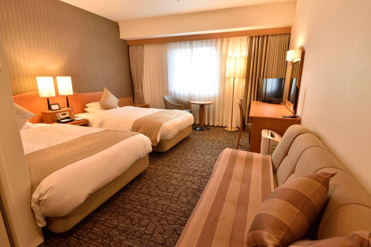 Quarto de hotel com duas camas, uma escrivaninha, uma cadeira, uma TV e janela com cortinas.