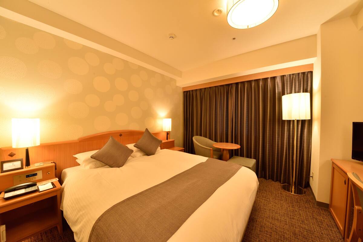 Quarto de hotel com uma cama grande, uma escrivaninha com telefone, uma cadeira, uma mesinha e uma televisão.