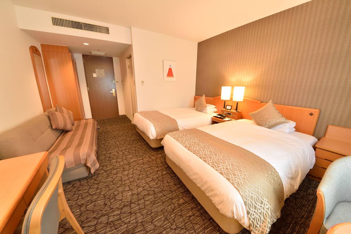 Quarto de hotel com duas camas, uma escrivaninha, uma cadeira e uma mesinha.