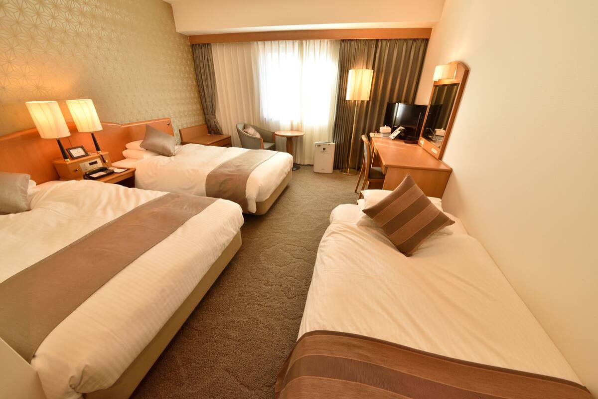 Quarto de hotel com duas camas, uma escrivaninha, uma cadeira, um abajur e um espelho.