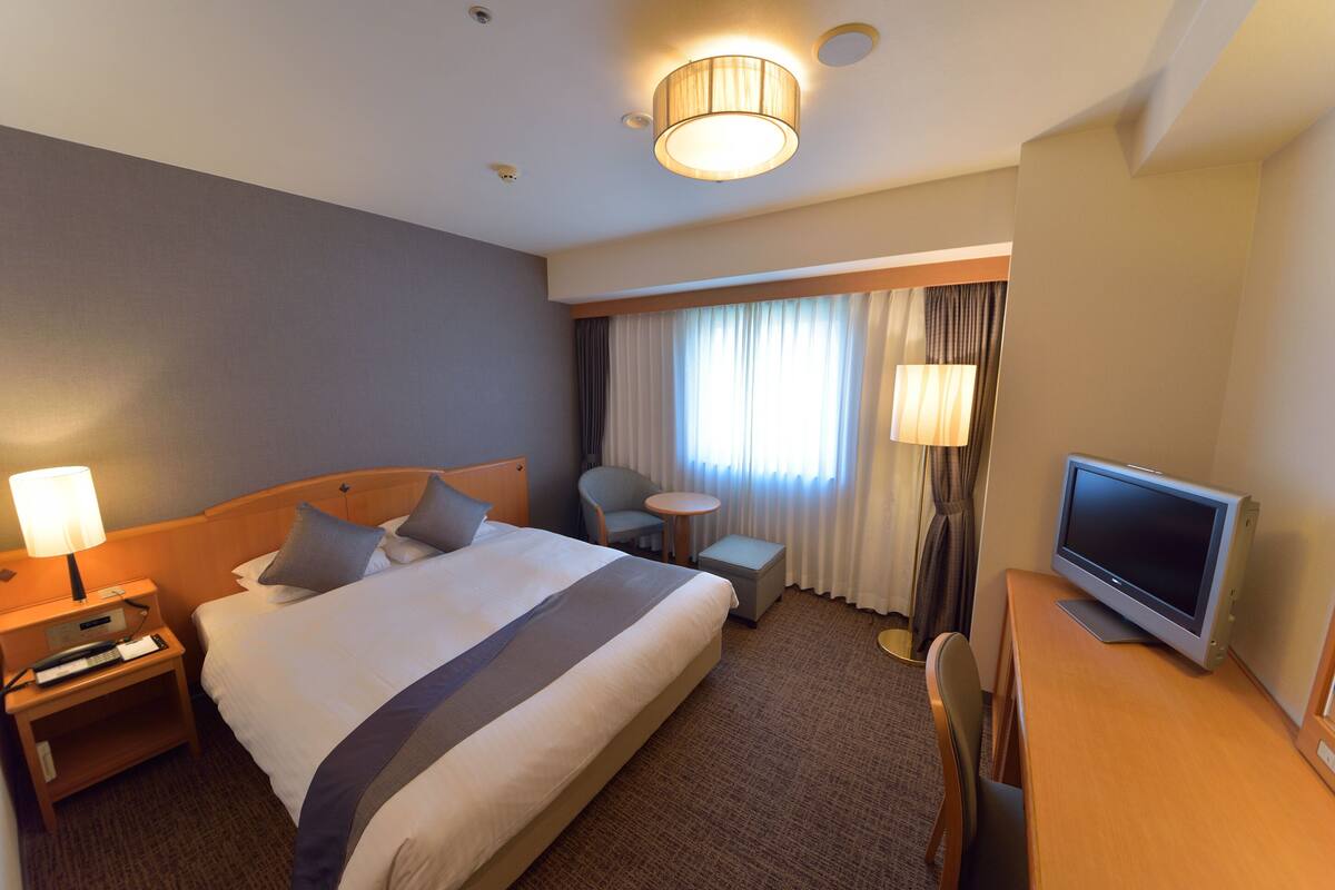 Quarto de hotel com uma cama grande, mesa de cabeceira com abajur, televisão, uma pequena mesa redonda e uma cadeira.