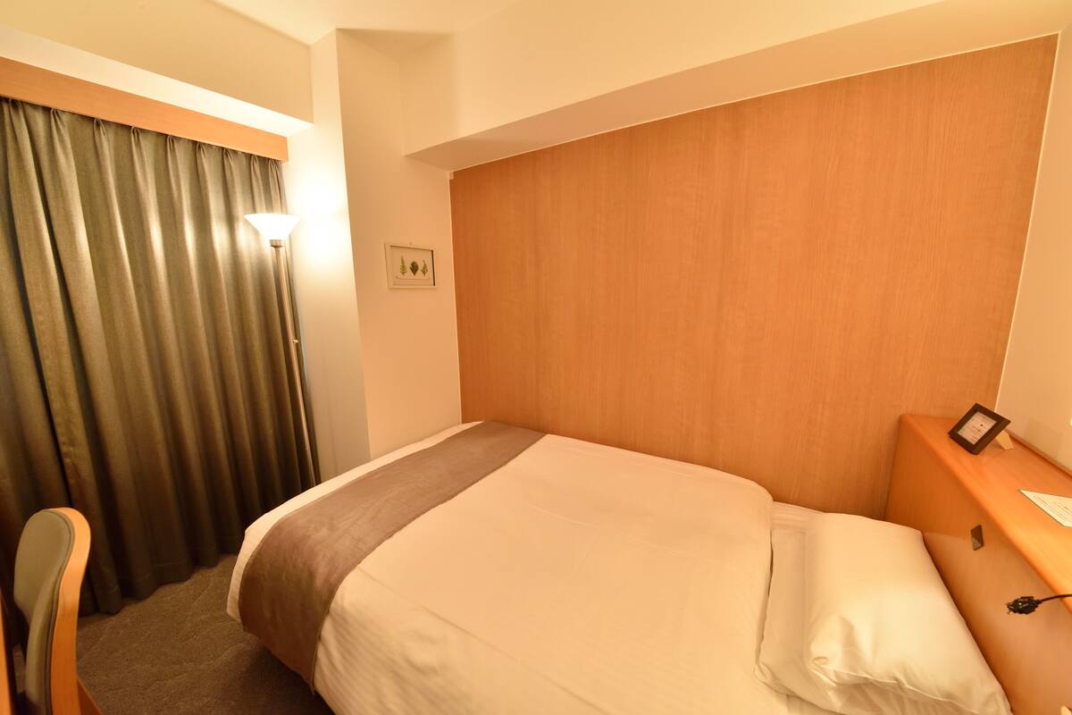 Quarto de hotel com cama, escrivaninha, cadeira, abajur e janela com cortinas.