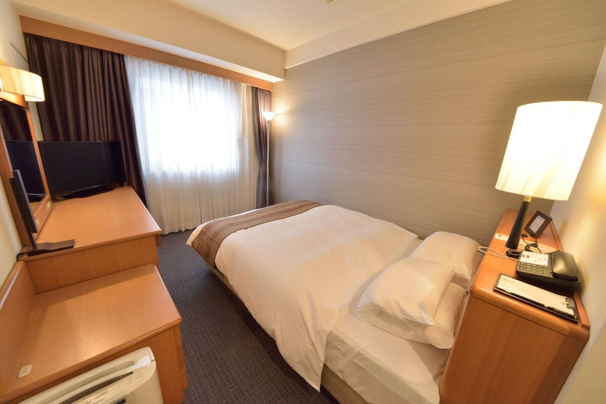 Quarto de hotel com cama, escrivaninha, cadeira, televisão, abajur e telefone.