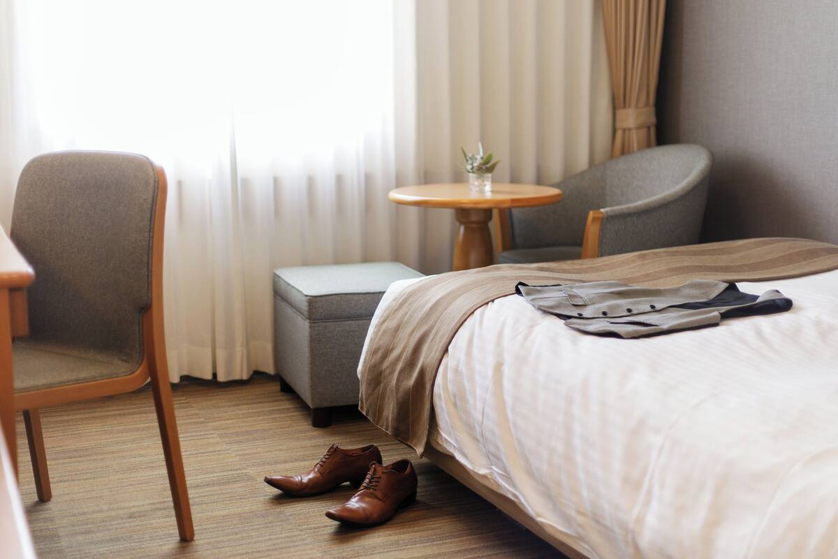Quarto de hotel com cama, uma cadeira, uma mesinha e um criado-mudo.