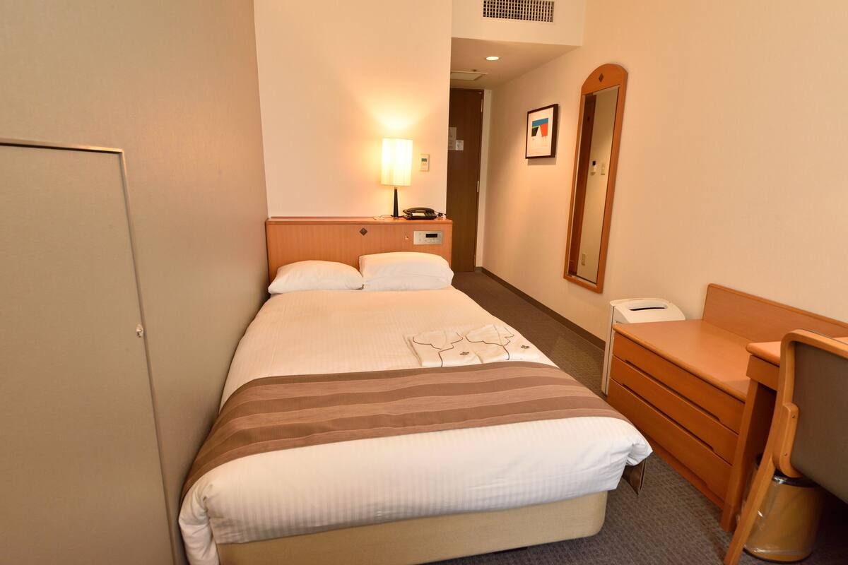 Quarto de hotel com cama, mesa de cabeceira, abajur, espelho e cômoda.