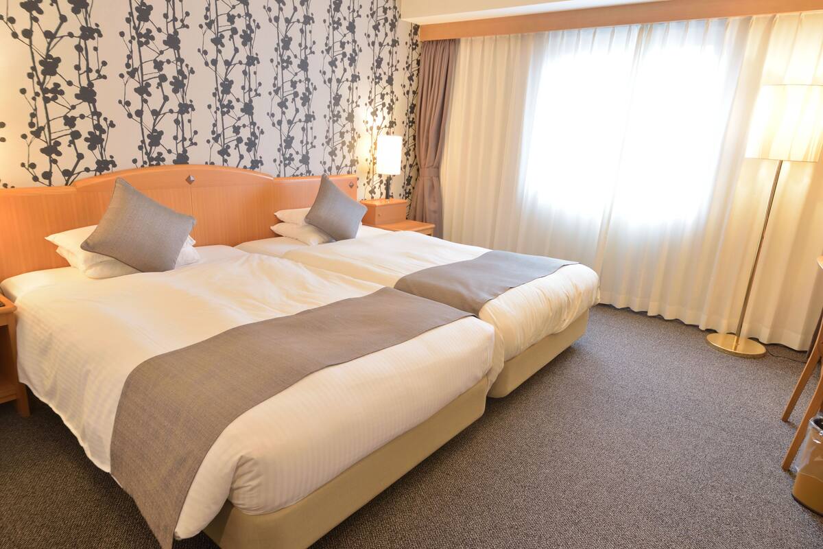 Uma cama de casal com duas almofadas, uma mesa de cabeceira, uma luminária e uma janela com cortinas.