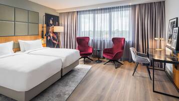 Quarto de hotel com uma cama grande, duas poltronas vermelhas, uma mesinha e uma escrivaninha com cadeira.