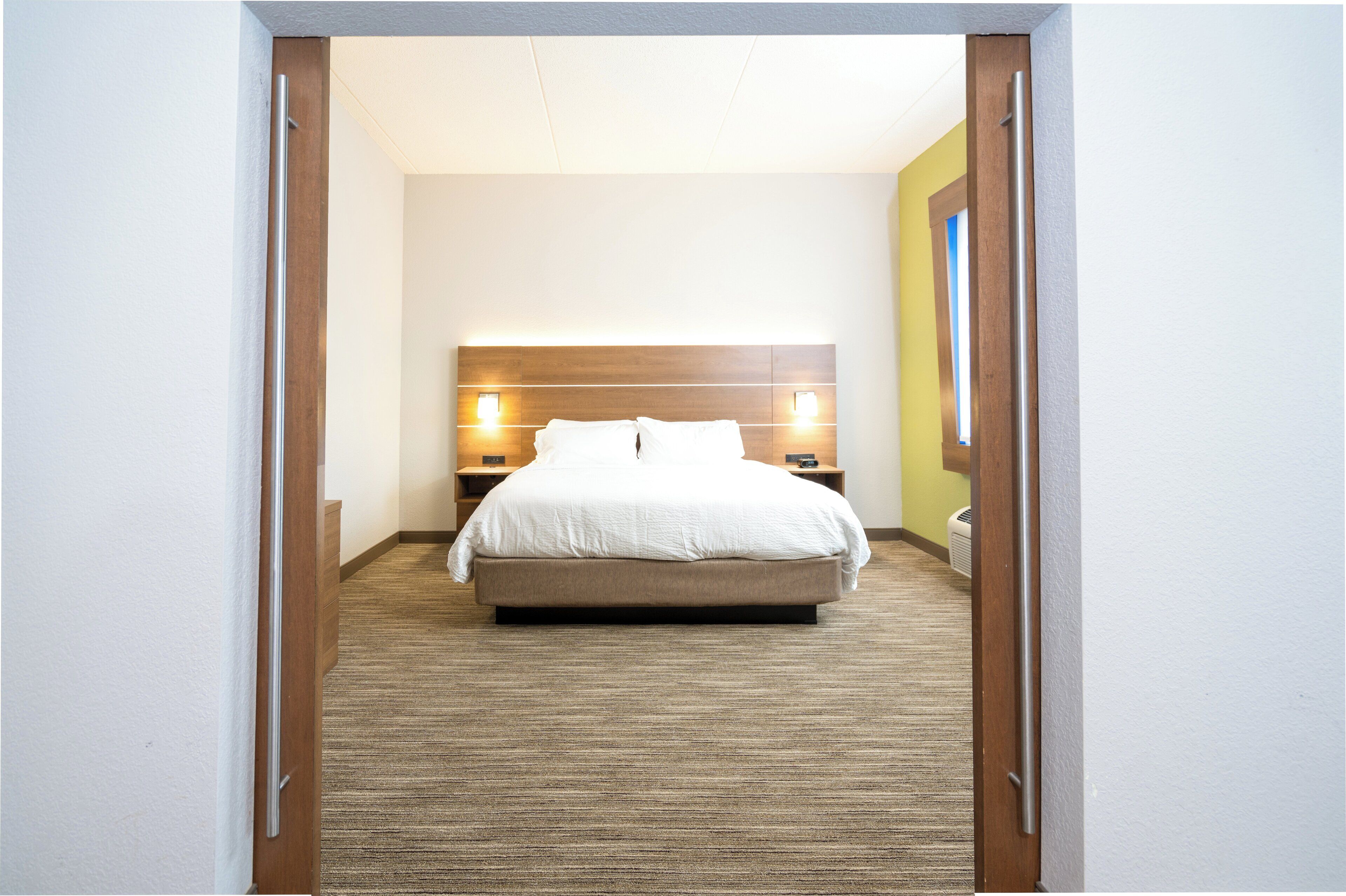 Quarto de hotel com uma cama grande, duas luminárias de cabeceira, uma cabeceira de madeira e piso coberto por carpete.