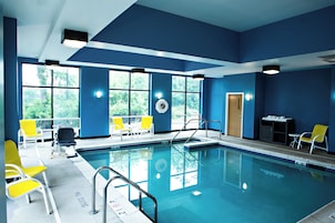 Piscina interior com água azul cristalina, cadeiras de descanso amarelas e brancas, e grandes janelas com vista para o verde lá fora.