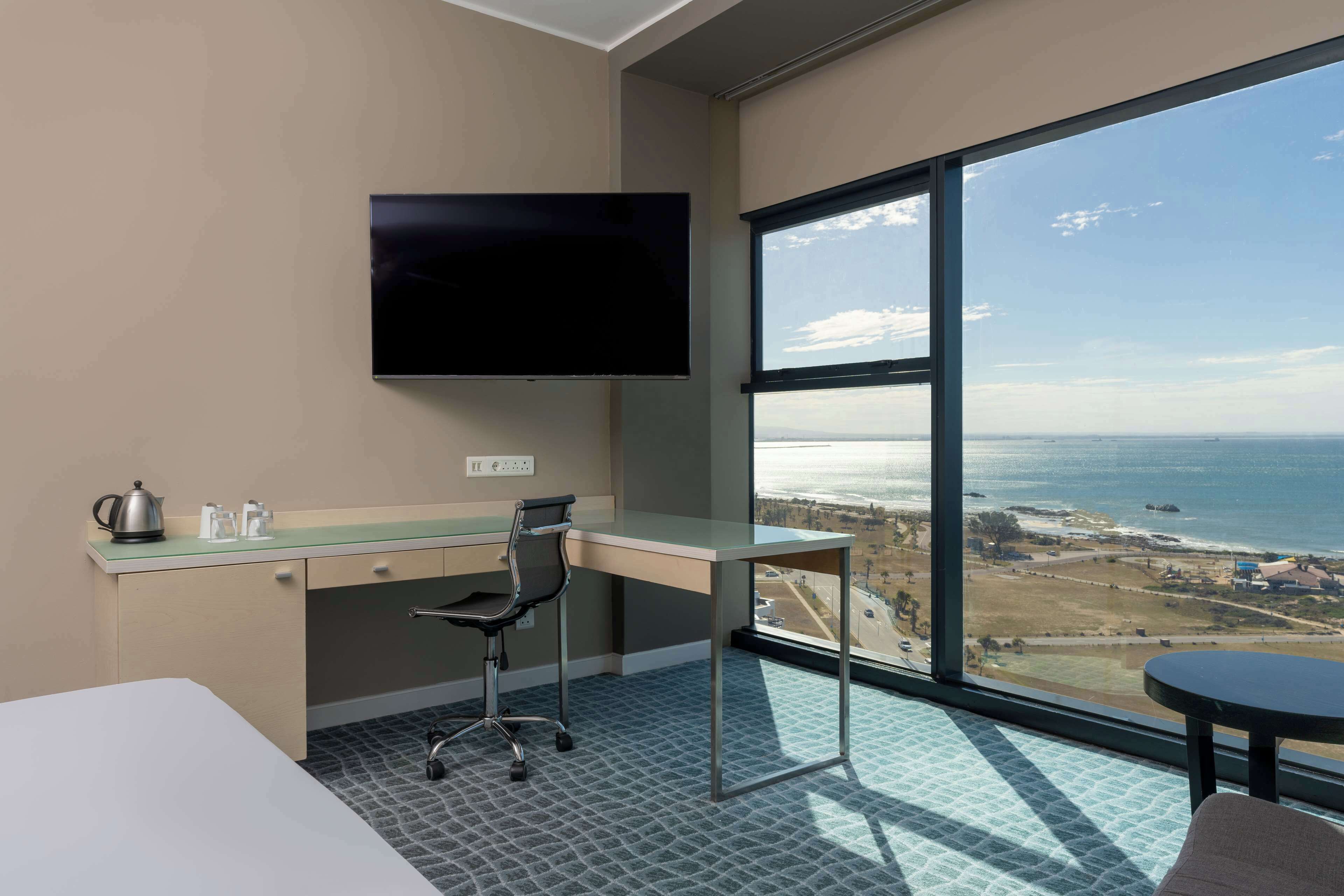 Quarto de hotel com uma grande janela que oferece vista para o mar e a praia, uma mesa com cadeira, televisão de tela plana e uma pequena mesa.