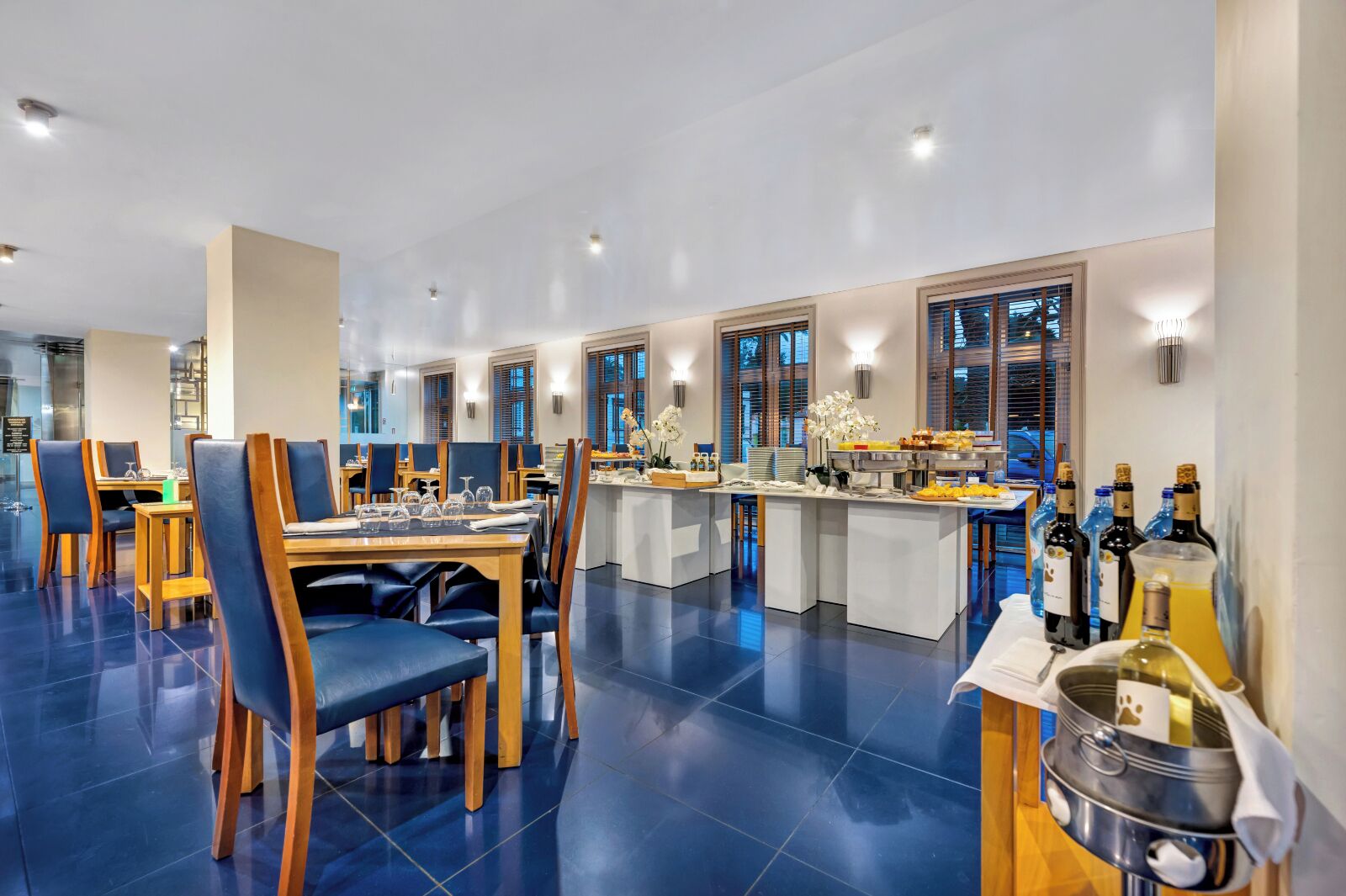Uma área de refeições moderna em hotel, com várias mesas e cadeiras, um buffet e um bar com garrafas de vinho e uma ponche.