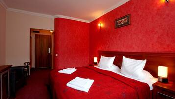 Quarto de hotel com carpete vermelho, cabeceira de madeira, cama com lençóis brancos, duas luminárias de mesa e um quadro pendurado na parede.