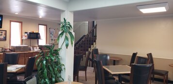 Uma sala de jantar com mesas e cadeiras de madeira, uma planta em vaso e uma cozinha com eletrodomésticos e geladeira.