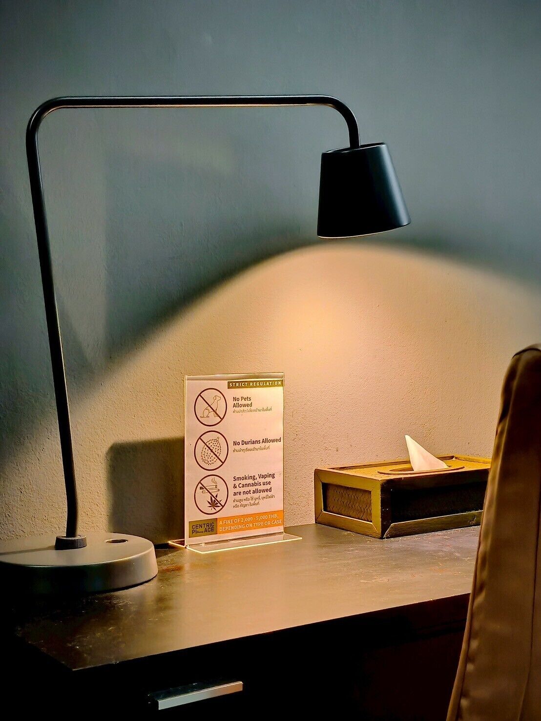 Uma mesa com uma lâmpada, uma caixa de lenços e um aviso com regulamentos rígidos, incluindo a proibição de animais de estimação, duriãs, fumar, usar cigarros eletrônicos ou cannabis.