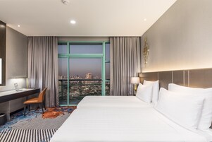 Quarto de hotel com uma cama grande, uma escrivaninha e uma cadeira. Vê-se a cidade através de uma janela ampla.
